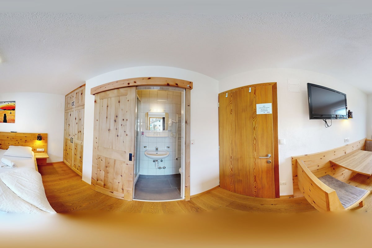 Quarto com cama, guarda-roupa de madeira, banheiro e televisão presa na parede.