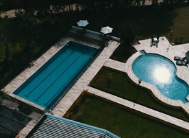 Vista aérea de uma área de piscina com uma piscina retangular e uma banheira de hidromassagem circular, cercada por um deck com assentos e guarda-sóis.