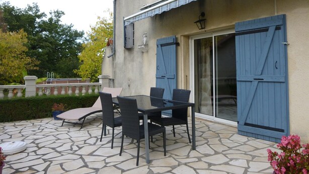 Um pátio externo com piso de pedra, uma mesa com cadeiras e persianas azuis no edifício.
