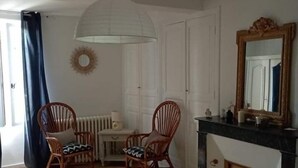 Quarto com uma cama, duas cadeiras de madeira, uma mesinha, radiador, espelho e janela com cortinas.