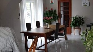 Uma área de refeições com mesa e cadeiras de madeira, uma grande planta em vaso e vista para o exterior.