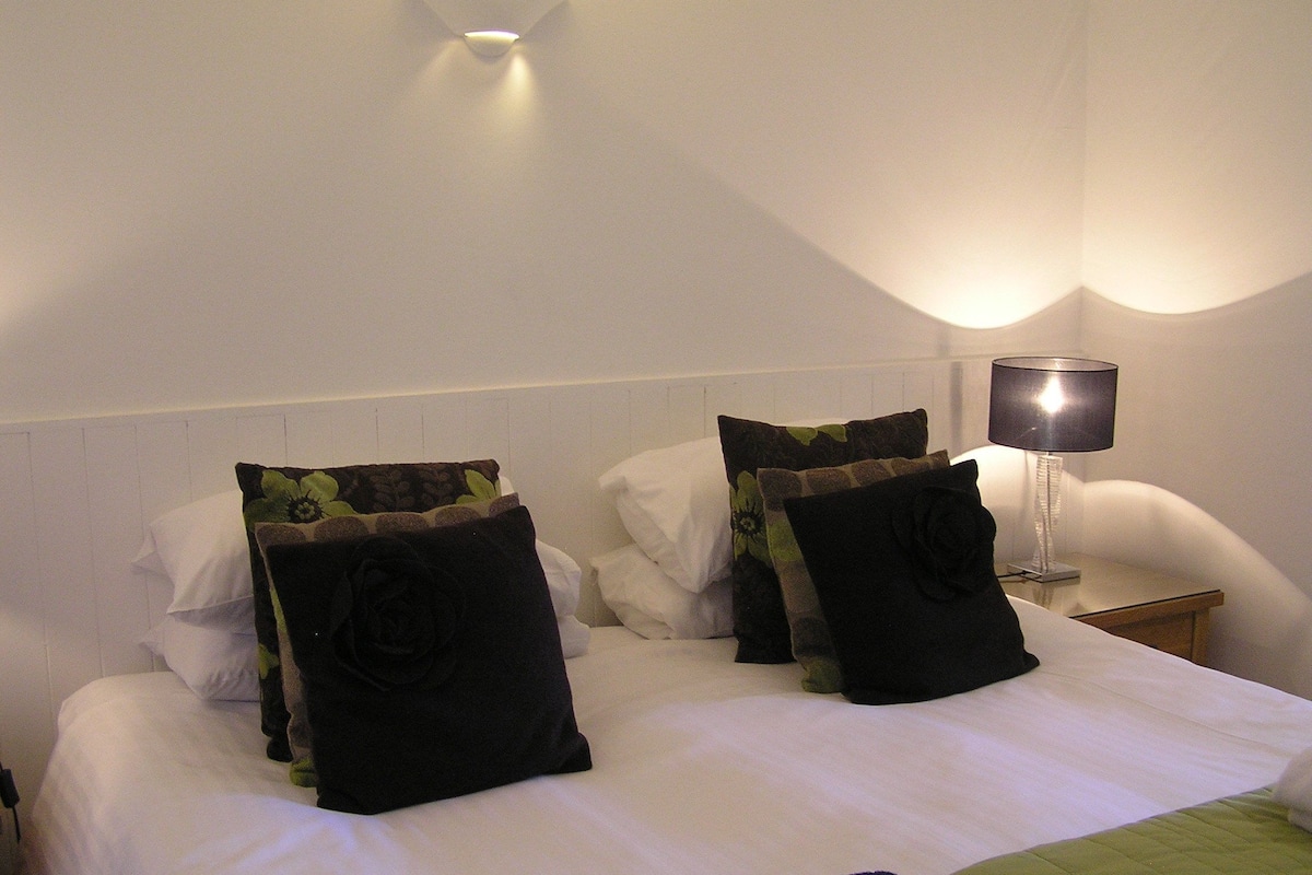 Quarto de hotel com cama, almofadas, abajur e luminária na parede.