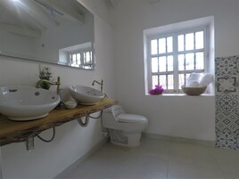Casa de banho com duas pias, sanita, janela e espelho, com uma cesta de toalhas.