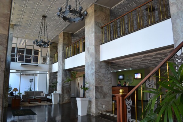 Um hall amplo com colunas de mármore, um balcão de recepção e uma escadaria com grades decorativas.