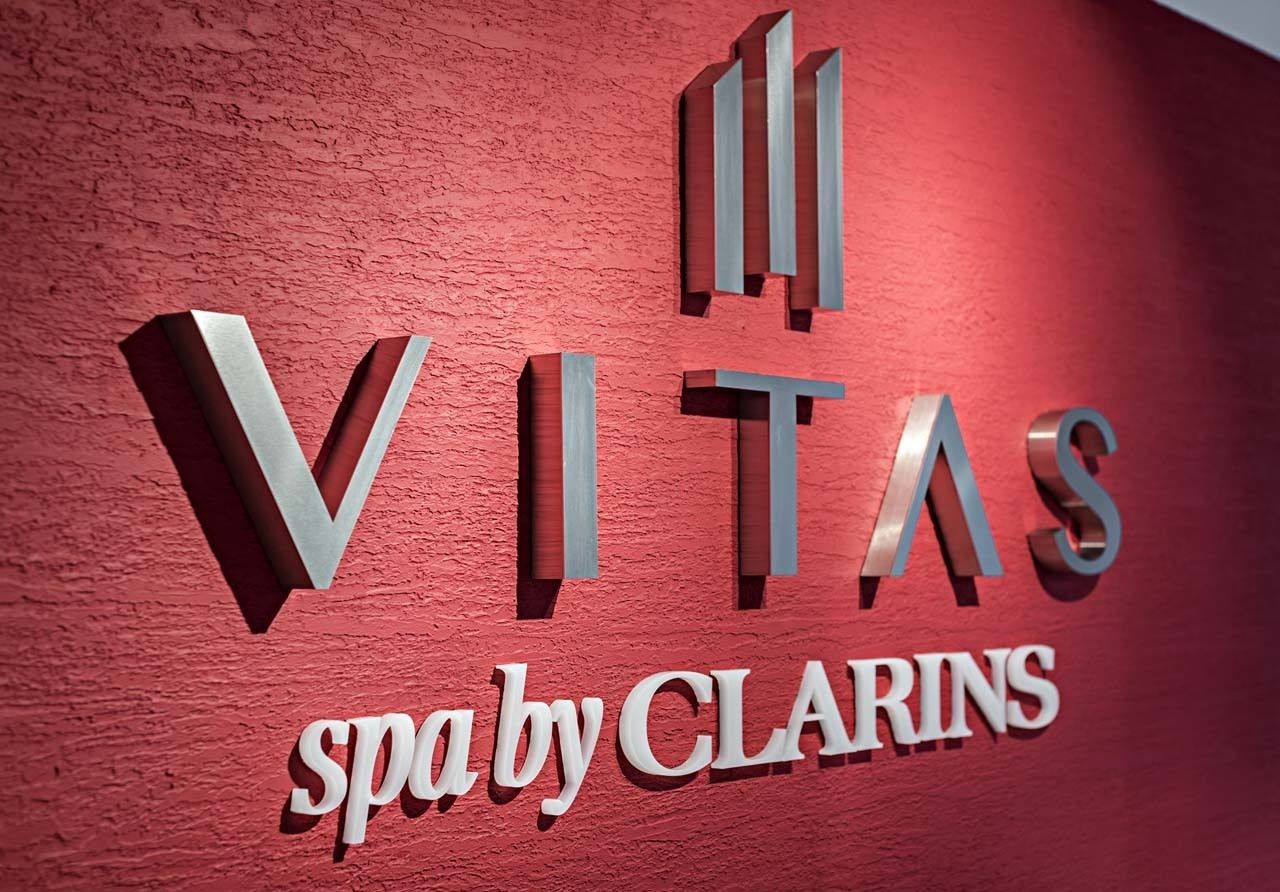 Um sinal 3D com o texto 'VITAS spa by CLARINS' em uma parede vermelha texturizada.