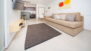 Uma sala de estar moderna com um sofá bege, uma TV de tela plana na parede e uma cozinha ao fundo.
