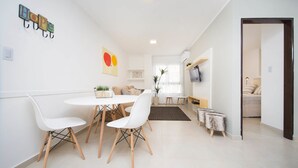 Um quarto moderno e bem iluminado, com área de jantar que inclui uma mesa redonda e cadeiras brancas. Há também um pequeno espaço de estar com um sofá e uma televisão fixada na parede.
