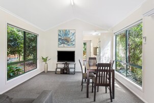 Uma sala de estar moderna com uma televisão de tela plana, uma mesa de jantar com cadeiras, janelas amplas com vista para áreas verdes e um quadro na parede.