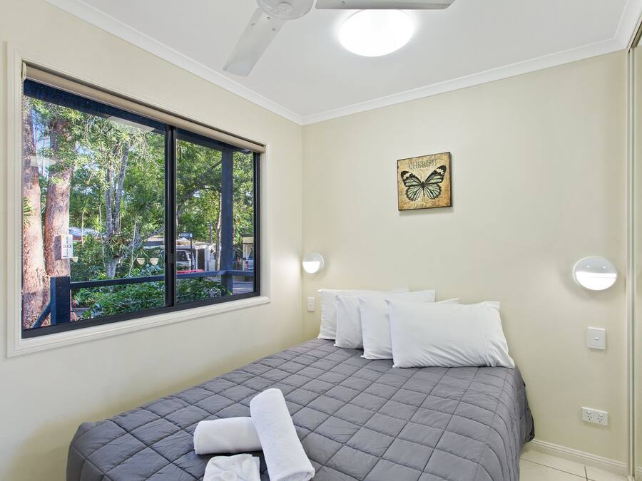 Quarto de hotel com uma janela grande, cama com colcha acolchoada cinza, almofadas brancas, uma obra de arte de borboleta na parede e um ventilador de teto.