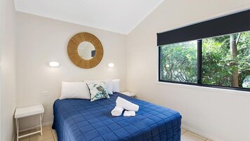 Um quarto com cama, mesa de cabeceira, espelho redondo e janela com vista para árvores.