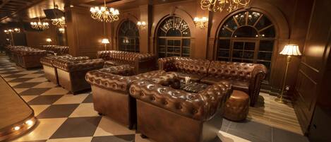 Um hall de entrada luxuoso com piso xadrez, sofás de couro e lustres ornamentados.