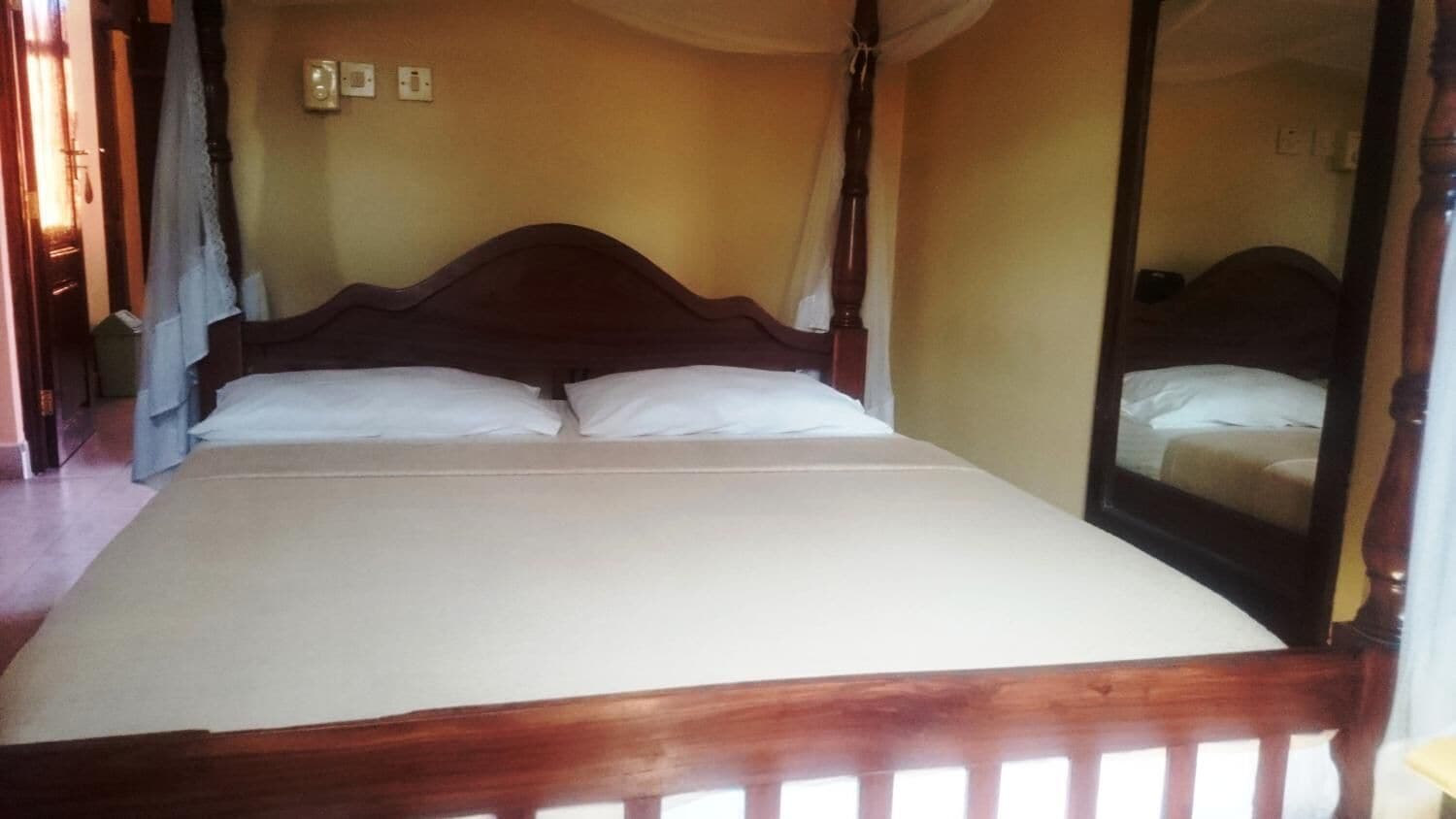 Uma cama grande com cabeceira de madeira e roupa de cama branca em um quarto com espelho e cortina.
