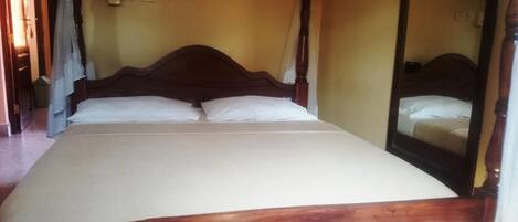 Uma cama grande com cabeceira de madeira e roupa de cama branca em um quarto com espelho e cortina.