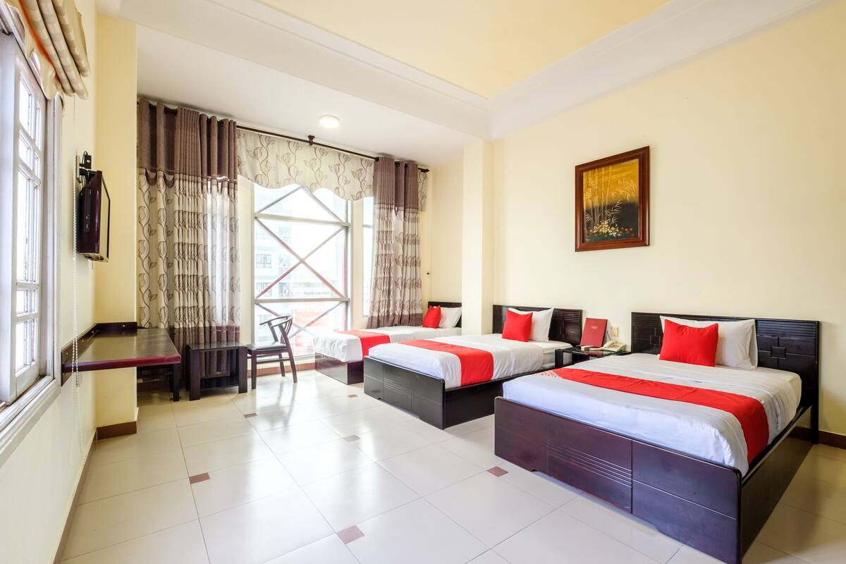 Quarto de hotel com duas camas, uma mesa e uma cadeira. Há uma janela com cortinas, um quadro na parede e vista para o exterior.