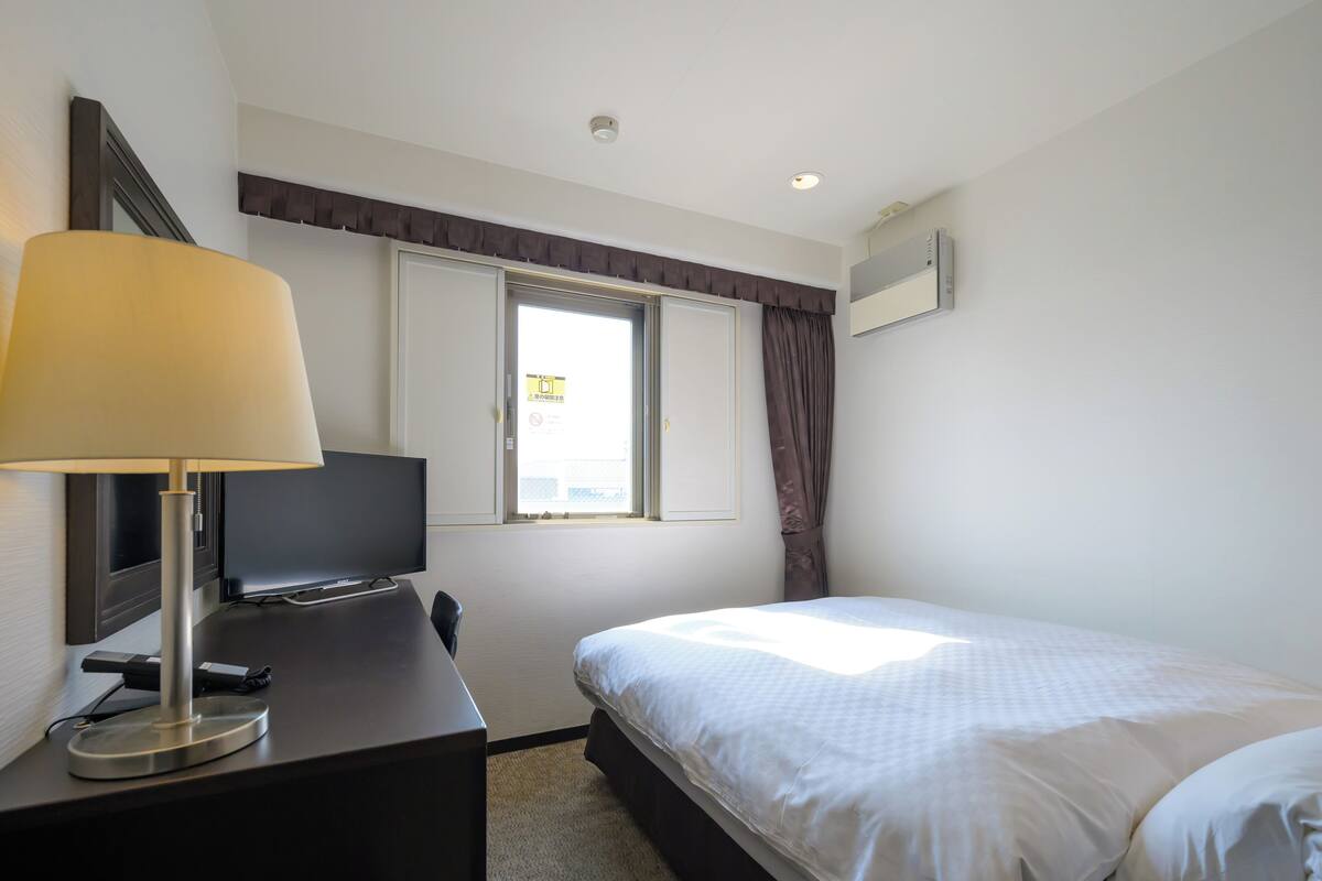 Quarto de hotel com cama, escrivaninha com luminária, televisão e janela com cortinas.