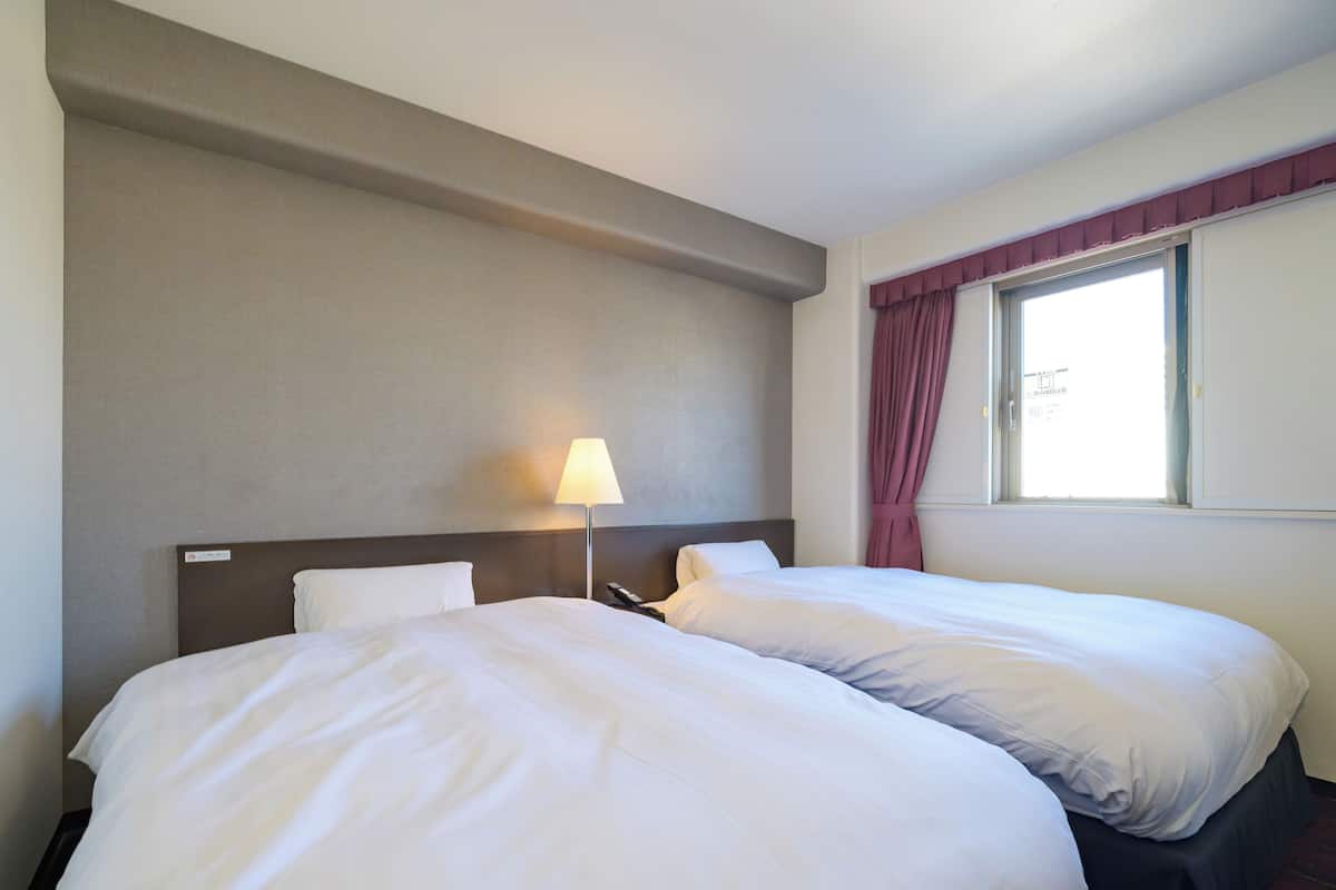 Quarto de hotel com duas camas, uma luminária de cabeceira e uma janela com cortinas.