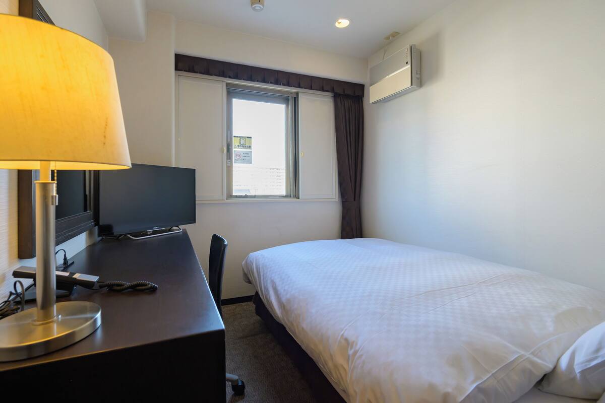 Quarto de hotel com escrivaninha, televisão, cama, janela com cortinas e aparelho de ar condicionado.