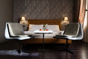 Quarto de hotel com uma cama, duas poltronas, uma mesa pequena com uma bandeja de comida e duas luminárias na parede.