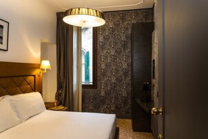 Quarto de hotel com cama, mesa de cabeceira, luminária, janela com cortinas, televisão fixada na parede e porta para o banheiro.