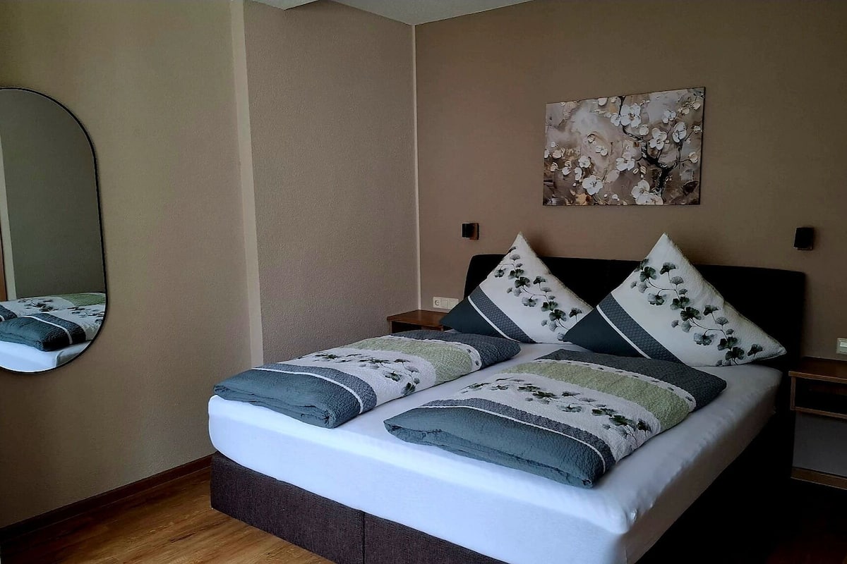 Uma cama bem arrumada com almofadas florais, uma mesa de cabeceira, um espelho fixado na parede e um quadro acima da cama.