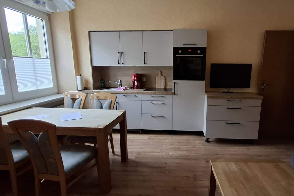Uma cozinha compacta com armários brancos, forno embutido e micro-ondas. Há uma mesa de jantar com cadeiras e um pequeno espaço de estar com um sofá.