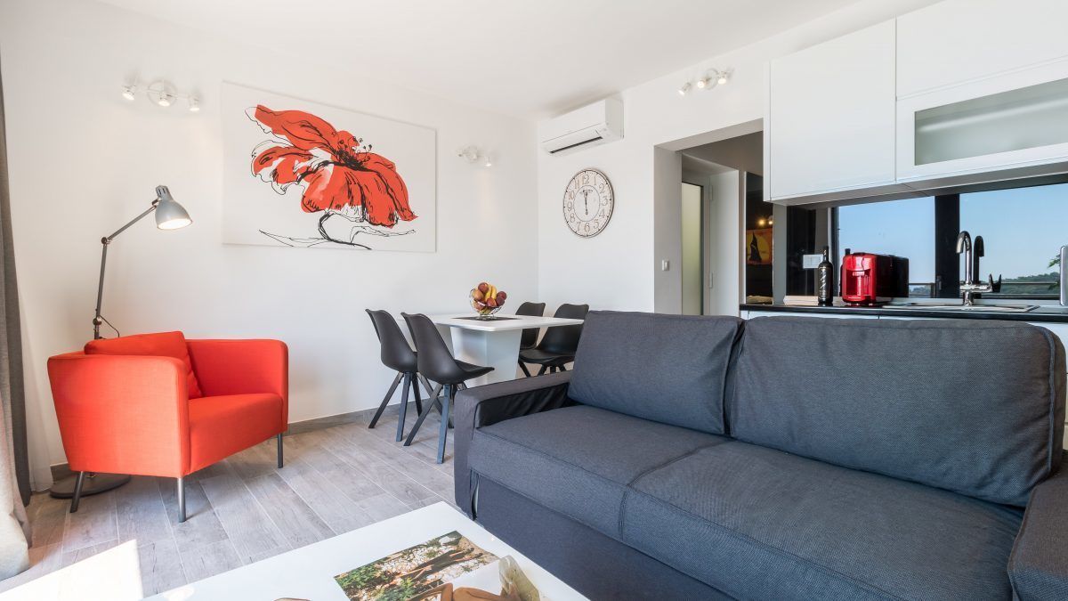 Uma sala de estar moderna com um sofá cinza, uma poltrona vermelha, uma mesa de jantar com cadeiras e uma área de cozinha com uma cafeteira vermelha.