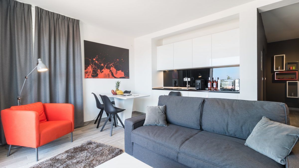 Uma sala de estar moderna com um sofá cinzento, uma poltrona vermelha e um espaço de refeições com mesa e cadeiras. Uma cozinha visível ao fundo apresenta armários brancos e uma janela.