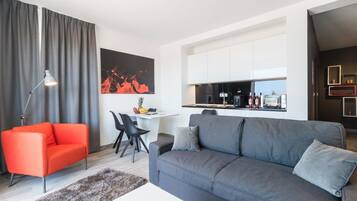 Uma sala de estar moderna com um sofá cinzento, uma poltrona vermelha e um espaço de refeições com mesa e cadeiras. Uma cozinha visível ao fundo apresenta armários brancos e uma janela.