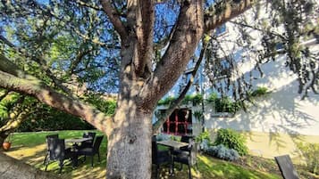 Uma área de estar externa com mesas e cadeiras sob uma grande árvore, um edifício branco com uma porta vermelha ao fundo e céu claro.