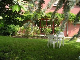 Um jardim com uma mesa e cadeiras de plástico branco, rodeado por vegetação exuberante e plantas floridas.