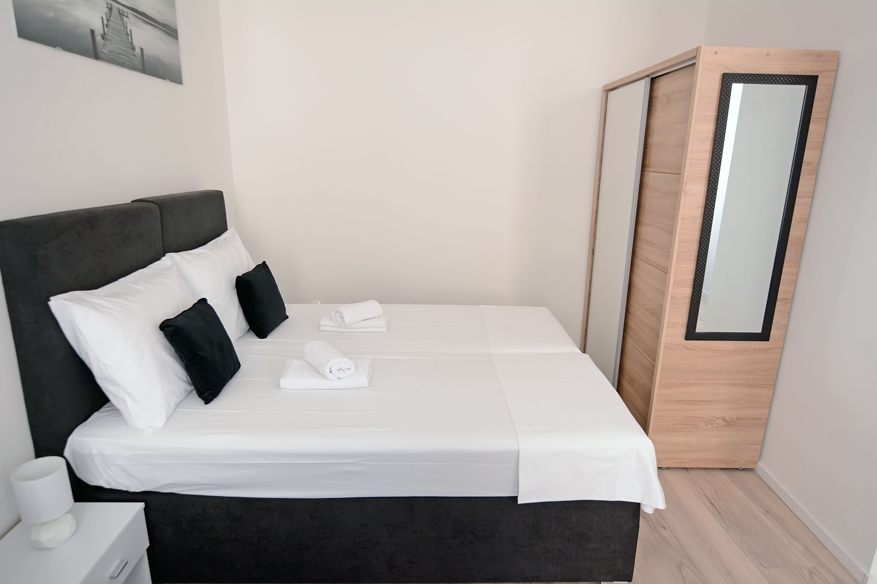 Cama bem arrumada com lençóis brancos e almofadas pretas, mesa de cabeceira com abajur branco e guarda-roupa de madeira com espelho.