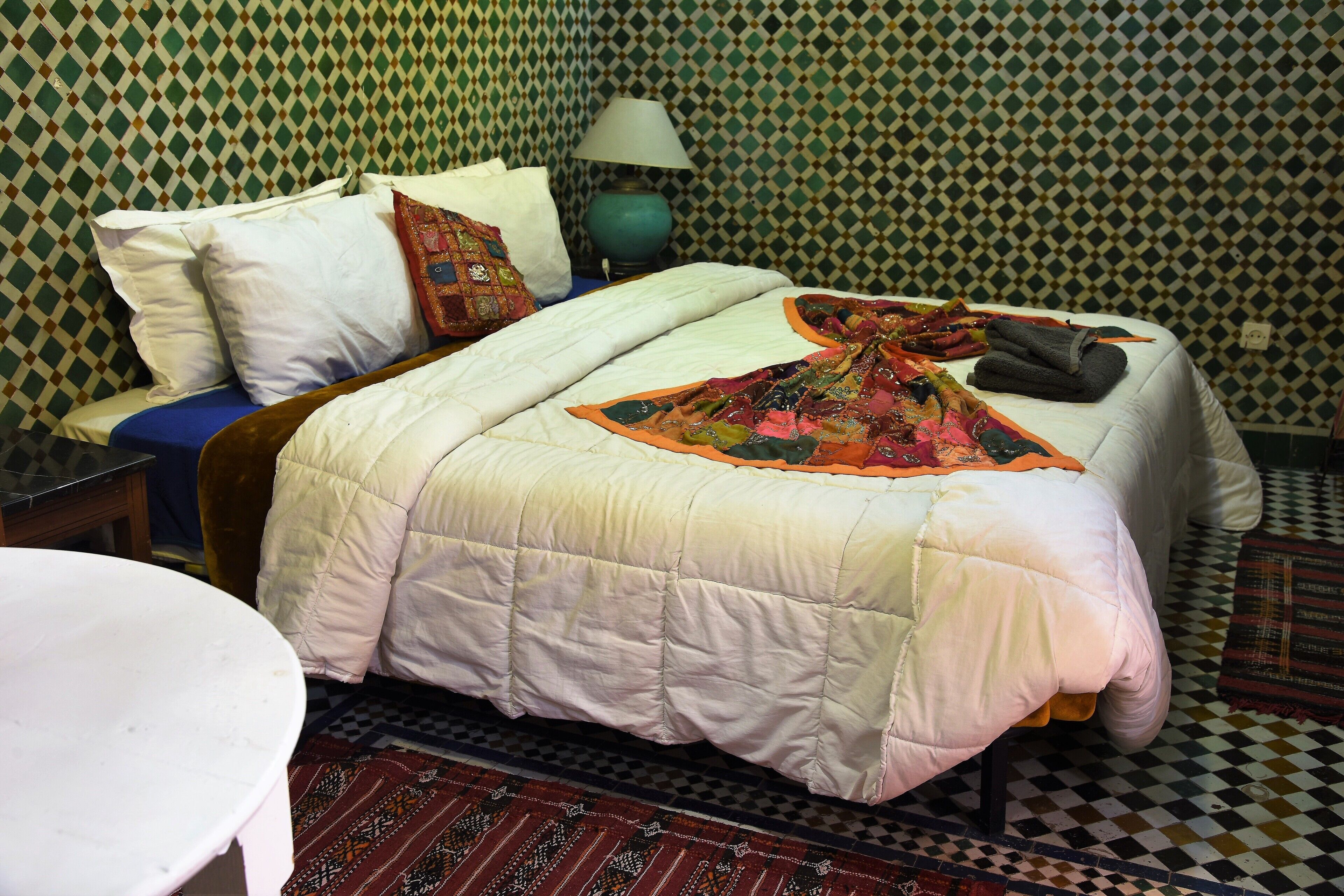Cama bem arrumada com colcha, travesseiros e cobertor. Há uma luminária em uma mesinha de cabeceira ao lado da cama. O quarto tem papel de parede estampado e chão xadrez.