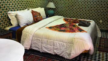 Cama bem arrumada com colcha, travesseiros e cobertor. Há uma luminária em uma mesinha de cabeceira ao lado da cama. O quarto tem papel de parede estampado e chão xadrez.