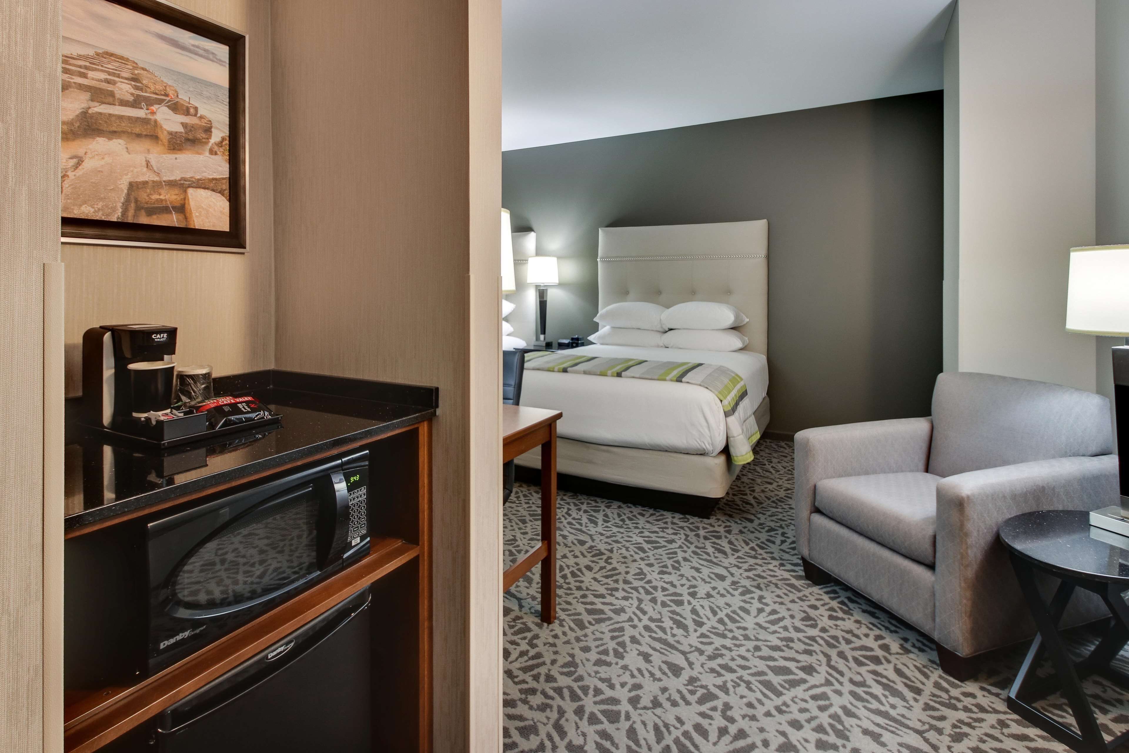 Quarto de hotel com cama, poltrona, escrivaninha, micro-ondas e máquina de café.