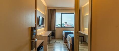 Quarto de hotel com escrivaninha, cadeira, duas camas, televisão e uma janela grande com vista para edifícios.