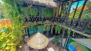 Uma área de piscina com um telhado de palha, rodeada por vegetação exuberante e uma ponte de madeira.