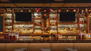 Um bar com parede de pedra ao fundo, prateleiras cheias de garrafas de bebidas e balcão com banquetas.