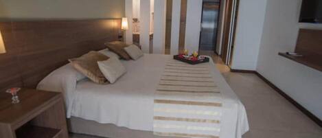 Quarto de hotel moderno com uma cama grande, mesas de cabeceira, televisão e um banheiro visível através de uma porta deslizante.