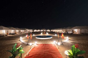 Diversas tendas dispostas em círculo, com uma piscina central, rodeadas por bandeiras e iluminadas por luzes.
