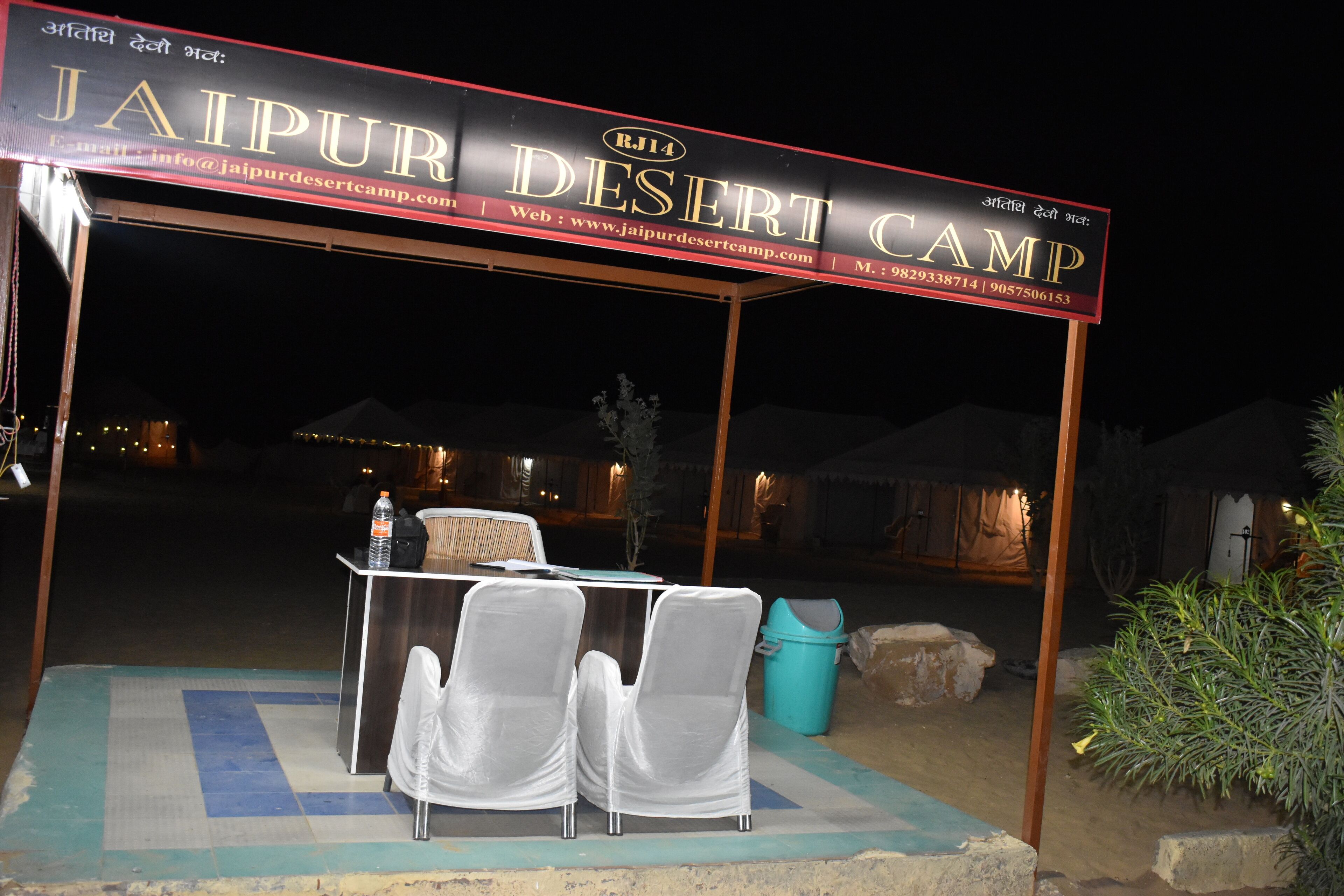 Acampamento no deserto com uma placa, uma mesa com cadeiras e um contentor de lixo.