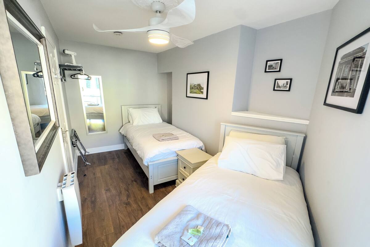 Quarto de hotel com duas camas de solteiro, um espelho grande, um ventilador de teto e quadros nas paredes.