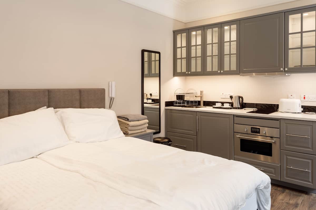 Quarto de hotel moderno com cama bem arrumada, pequena cozinha com cafeteira e armários cinzentos.