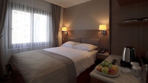 Quarto de hotel com uma cama grande, duas luminárias de cabeceira, mesa de cabeceira com telefone e máquina de café, e uma fruteira na mesinha lateral.