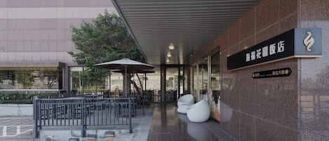 Entrada moderna de hotel com área de estar externa, galeria coberta e sinalização indicando a localização da recepção e do restaurante.