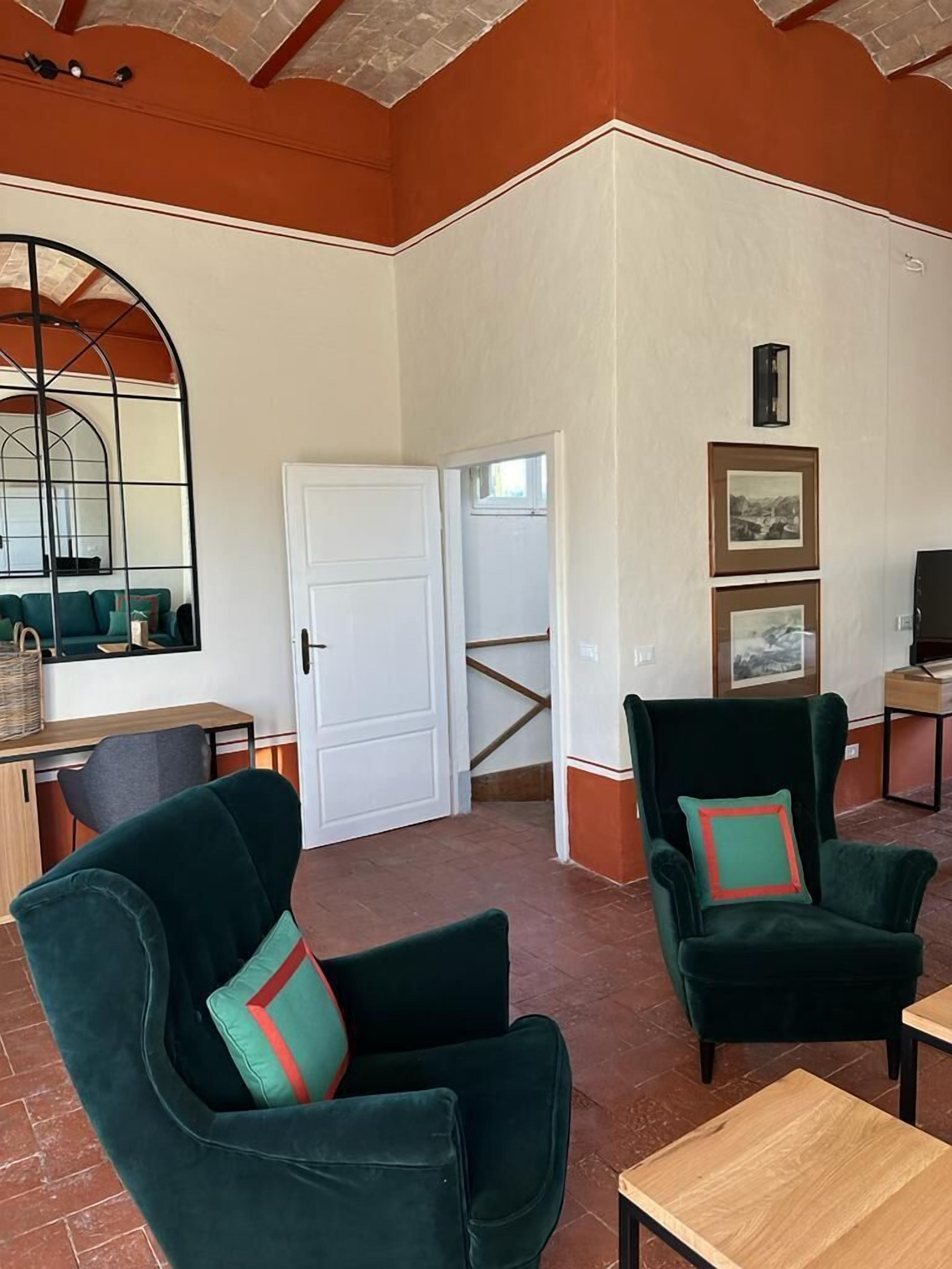 Quarto com duas poltronas verdes, uma mesa de madeira, uma porta branca e uma escada que leva a outro espaço.