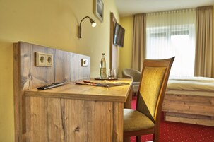 Quarto de hotel com escrivaninha de madeira, uma cadeira, uma cama, televisão e janela com cortinas.