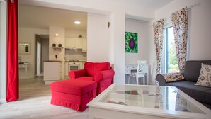 Uma sala de estar moderna com um sofá vermelho, uma mesa de centro de vidro e um sofá cinza com almofadas florais. Uma cozinha é visível ao fundo.