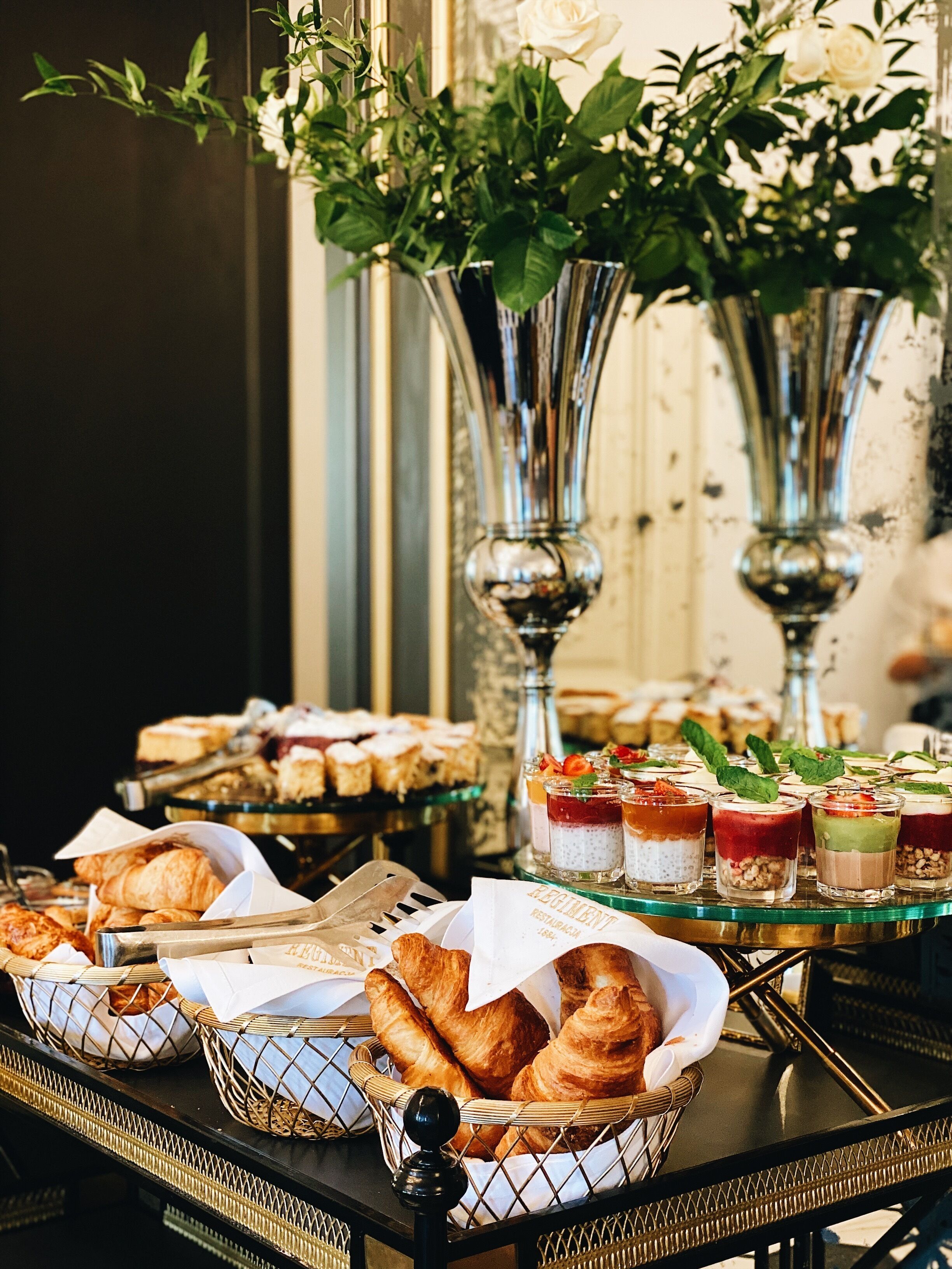 Uma mesa de buffet com diversos alimentos, incluindo doces e sobremesas, decorada com um vaso e um arranjo de flores ao fundo.