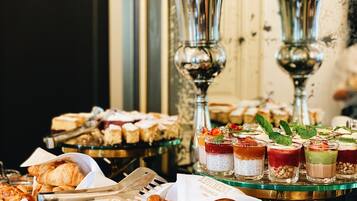 Uma mesa de buffet com diversos alimentos, incluindo doces e sobremesas, decorada com um vaso e um arranjo de flores ao fundo.