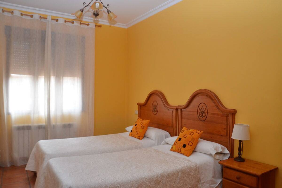 Quarto com duas camas, cabeceira de madeira, mesa de cabeceira com abajur e cortinas leves.